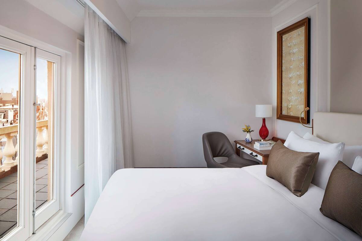 Quarto de hotel com uma cama grande, uma cadeira, uma escrivaninha com abajur, um quadro emoldurado e vista pela janela.