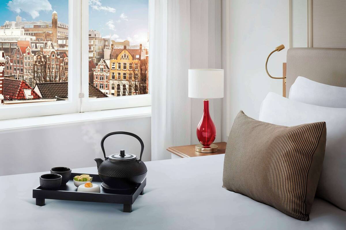 Quarto de hotel com cama, mesa de cabeceira com luminária, bandeja com chá e janela com vista para a cidade.
