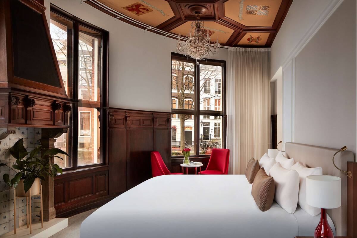 Quarto de hotel com uma cama grande, um candelabro, uma cadeira vermelha e vista para edifícios pela janela.