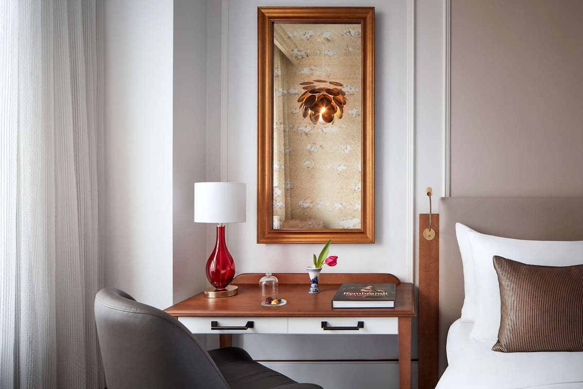 Quarto de hotel com cama, mesa de cabeceira com abajur, vaso com flor, quadro e um livro.