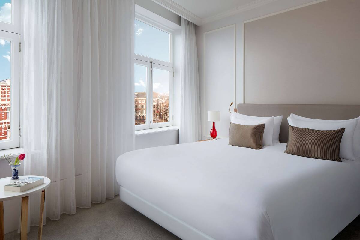 Quarto de hotel com uma cama grande, mesa de cabeceira, janela com cortinas e vista para edifícios.