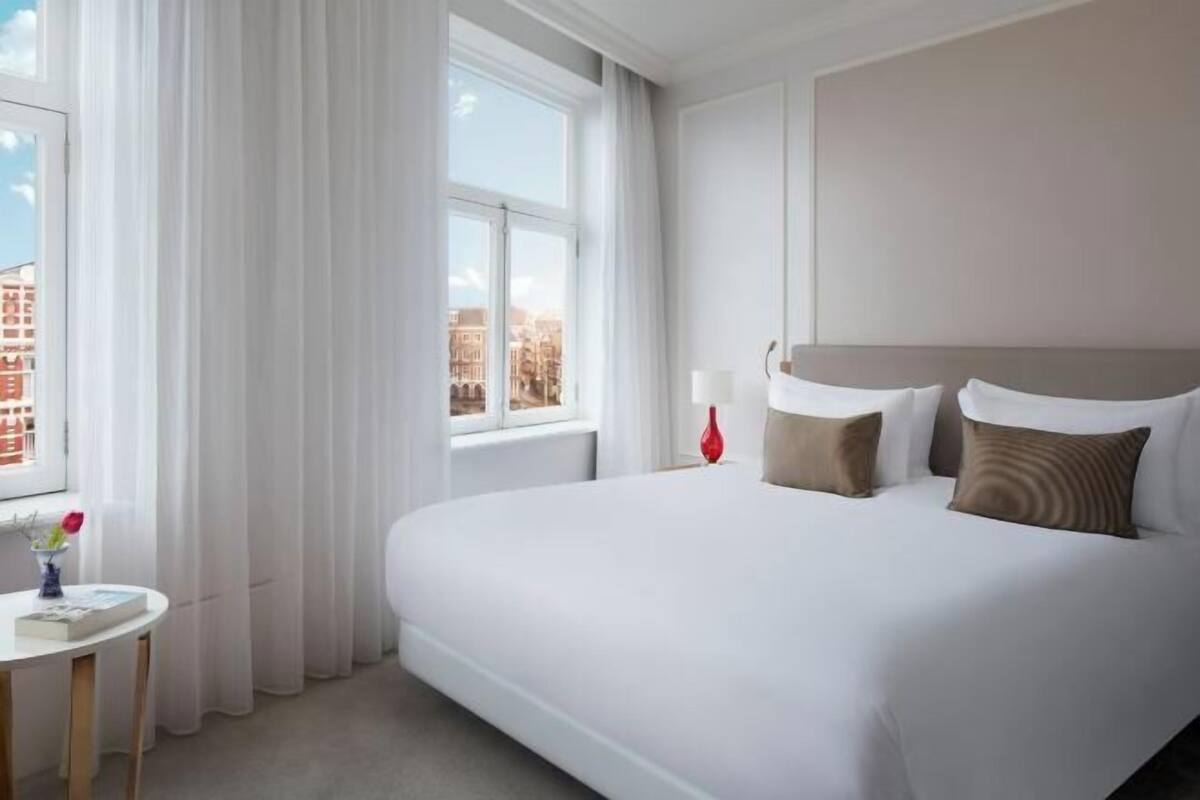 Quarto de hotel com uma cama grande, uma mesa pequena com um vaso e uma janela com cortinas.