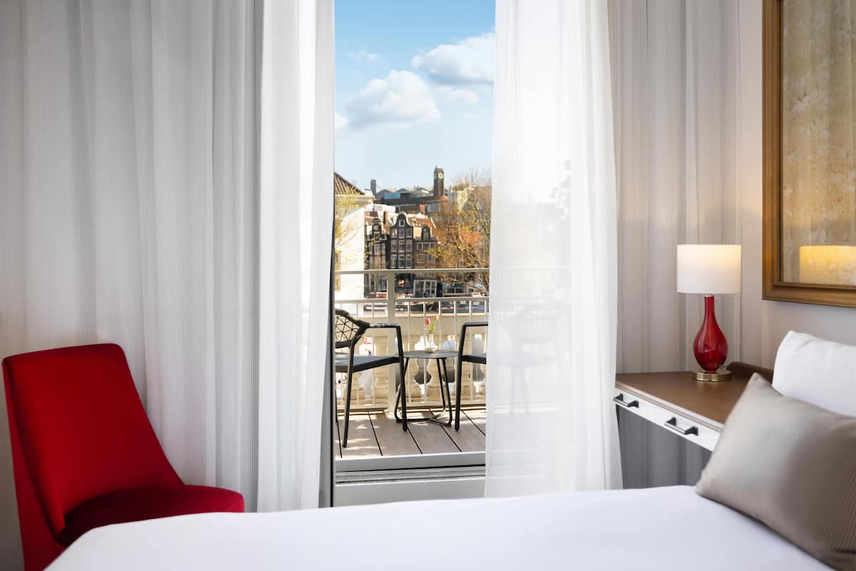 Quarto de hotel com cama, uma cadeira vermelha, mesa com abajur e vista para edifícios pela janela.