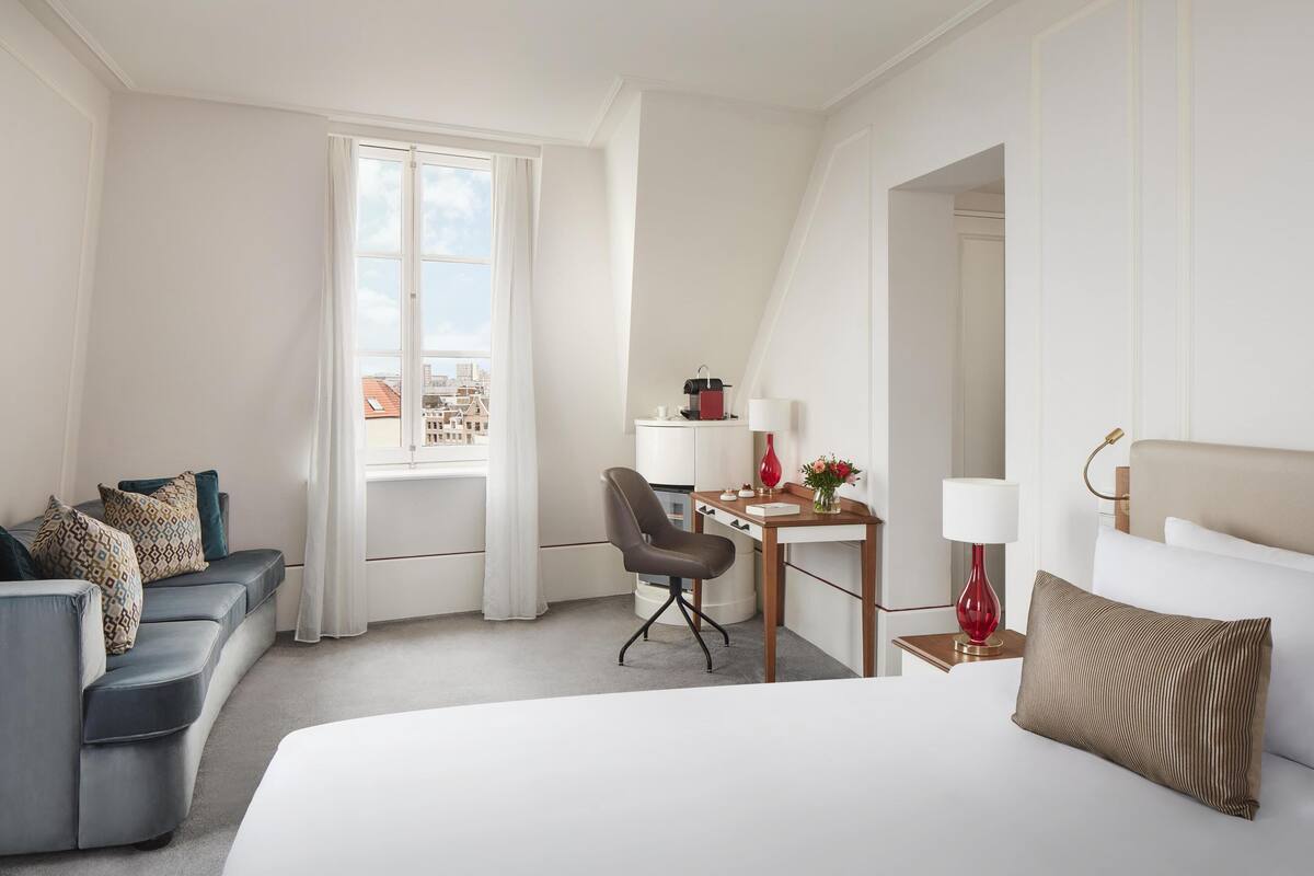 Quarto de hotel com cama, escrivaninha, cadeira, sofá, janela com cortinas e vista para a cidade.
