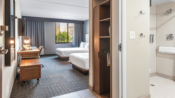 Quarto de hotel com escrivaninha, duas camas, guarda-roupa e área de chuveiro.