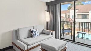 Quarto de hotel com sofá cinzento, um banquinho a combinar, uma janela grande com vista para uma piscina e edifícios, e um candeeiro de chão.