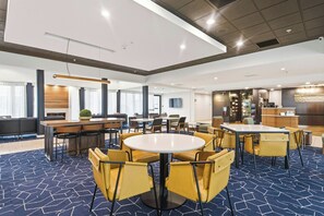 Um amplo hall de hotel com várias áreas de estar, incluindo mesas redondas e cadeiras, uma recepção e um bar.