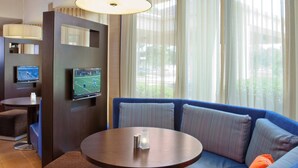 Quarto de hotel moderno com sofá azul, mesa redonda de madeira e televisão mostrando um jogo de tênis.