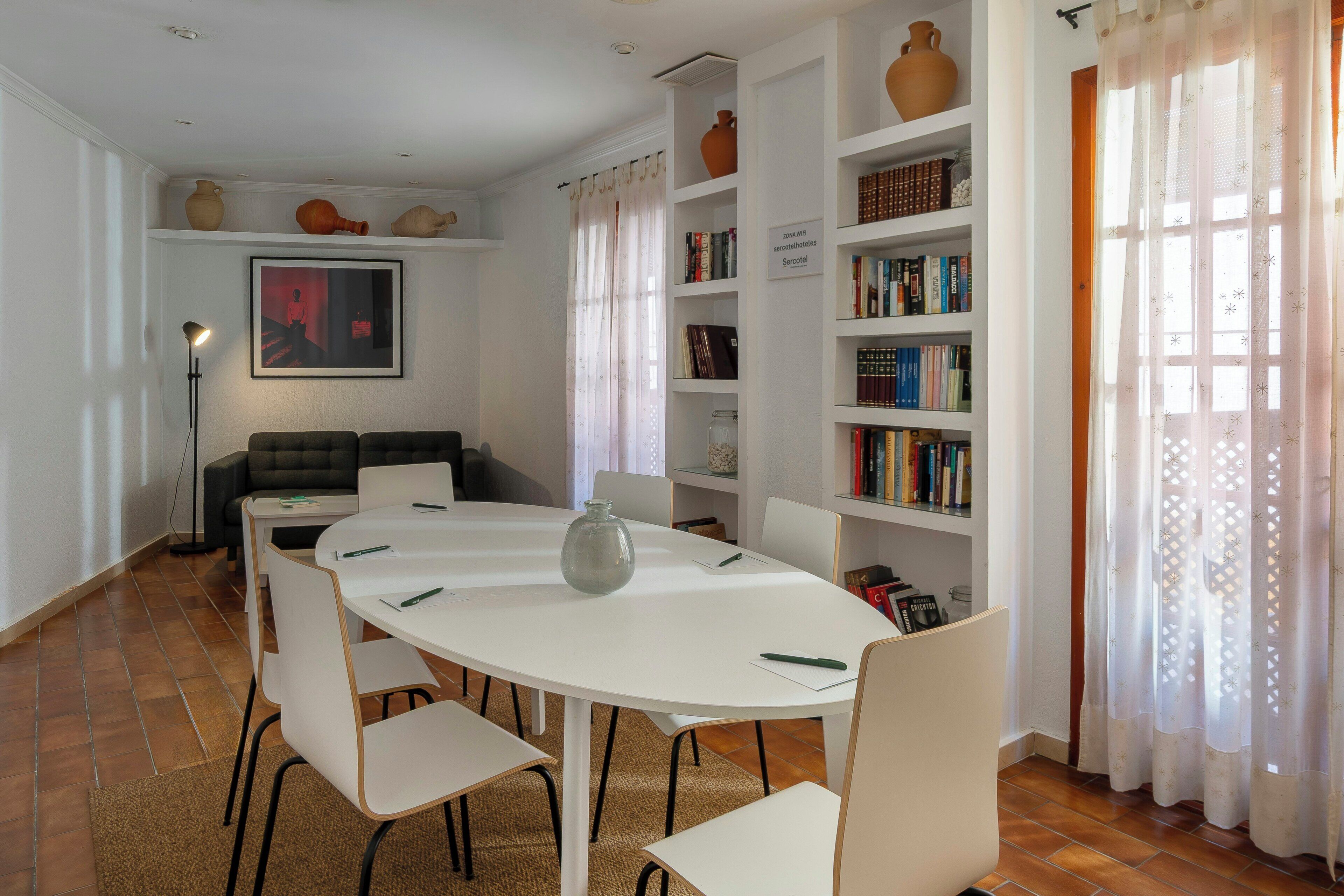 Uma sala de jantar com uma mesa branca, cadeiras, um vaso e uma estante com livros e objetos decorativos.