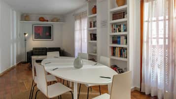 Uma sala de jantar com uma mesa branca, cadeiras, um vaso e uma estante com livros e objetos decorativos.
