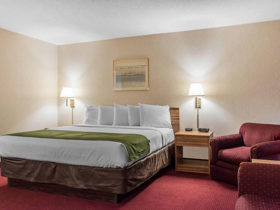 Quarto de hotel com uma cama grande, duas mesas de cabeceira com abajures, uma poltrona vermelha e um quadro na parede.