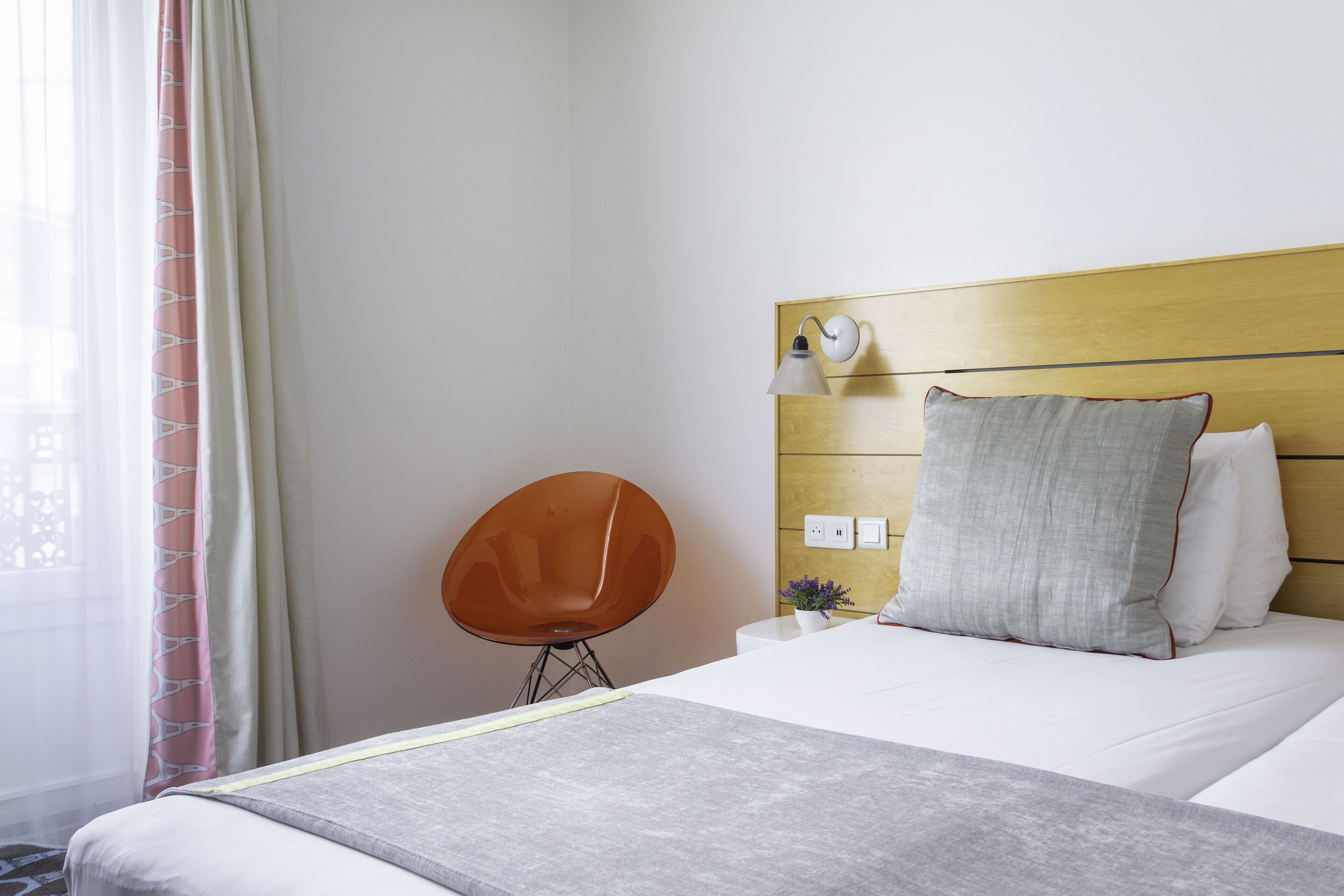 Quarto de hotel com cama, cabeceira de madeira, uma cadeira e janela com cortinas.