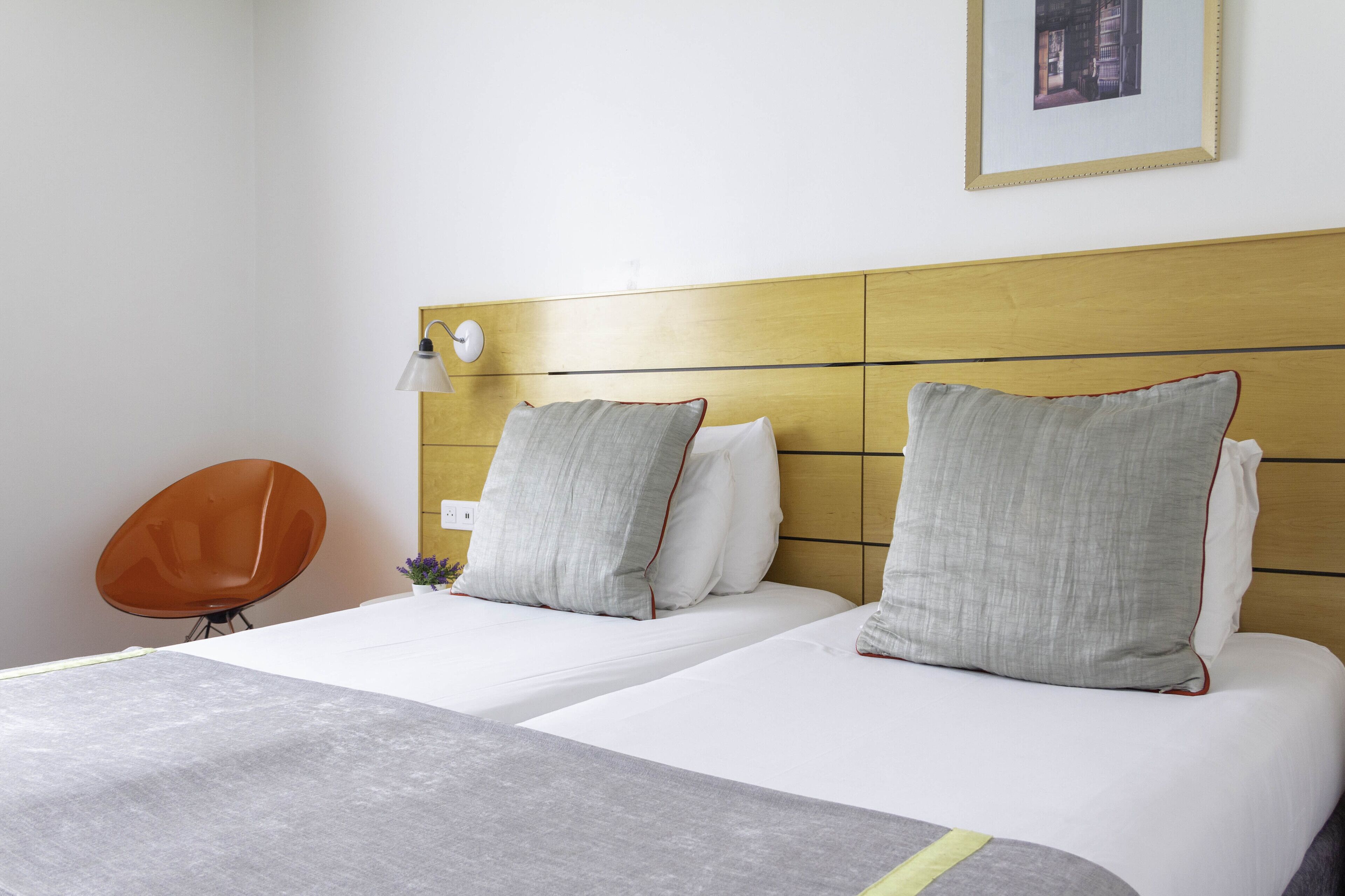 Quarto de hotel com duas camas, cabeceira de madeira, cadeira moderna e uma mesinha com uma planta.
