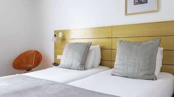 Quarto de hotel com duas camas, cabeceira de madeira, cadeira moderna e uma mesinha com uma planta.