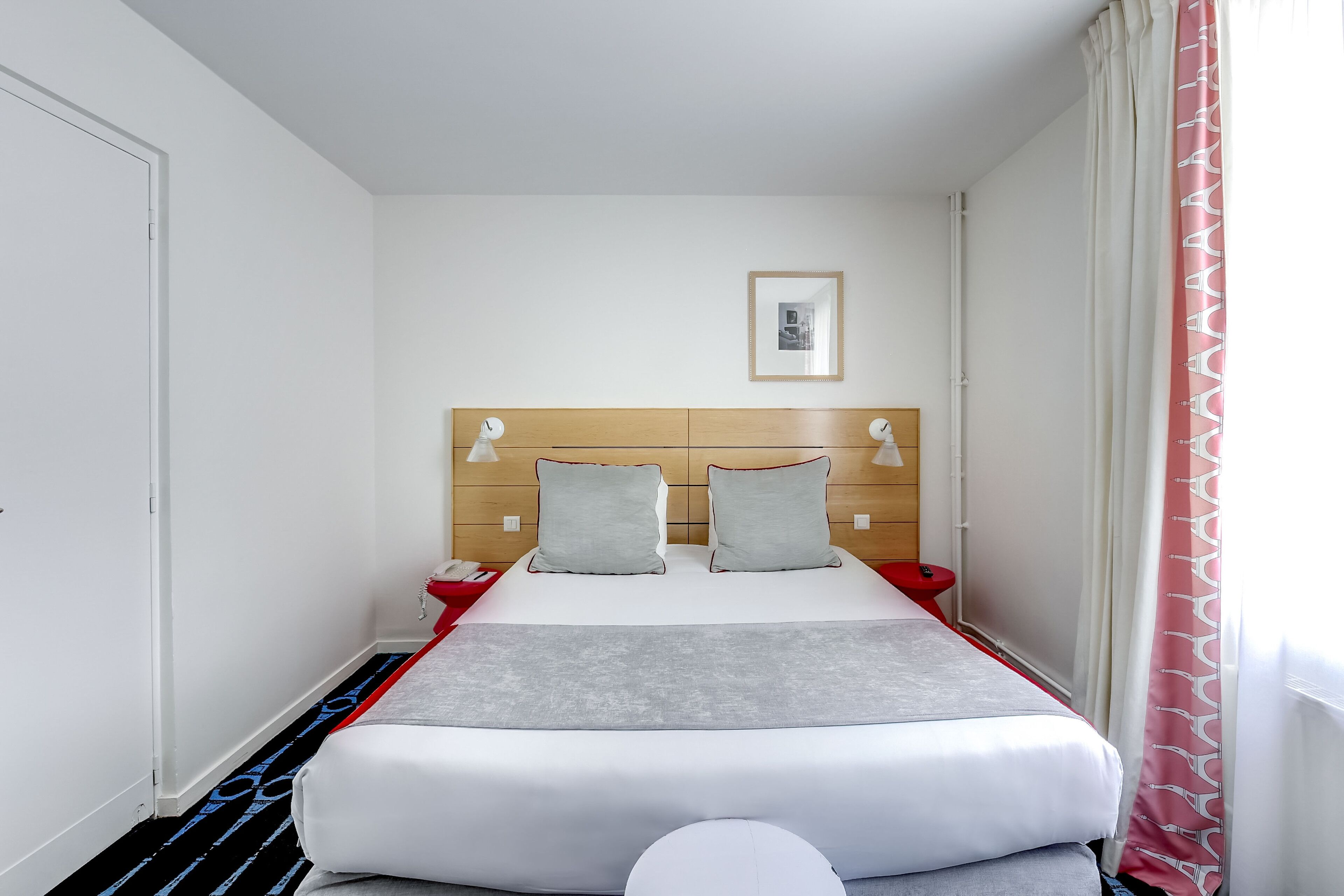 Quarto de hotel com cama, mesas de cabeceira, tapete vermelho, uma cadeira vermelha e uma janela com cortinas vermelhas.