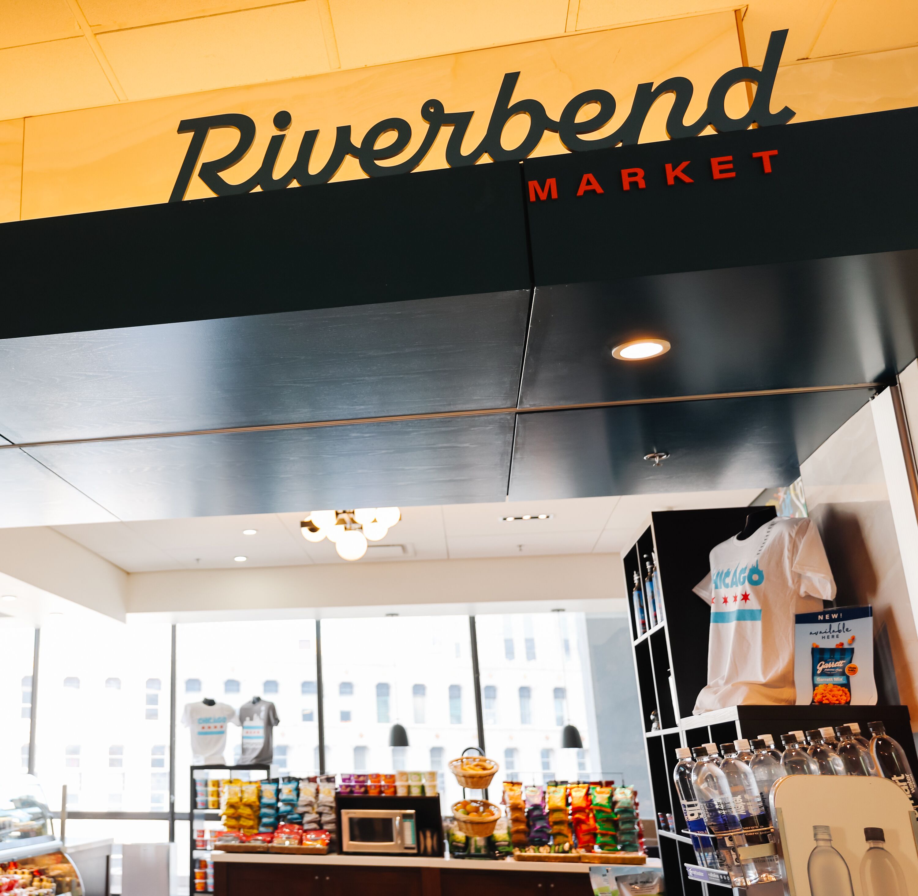 Fachada do Riverbend Market, com visão clara da entrada, diversos produtos embalados expostos e um aviso indicando novidades.