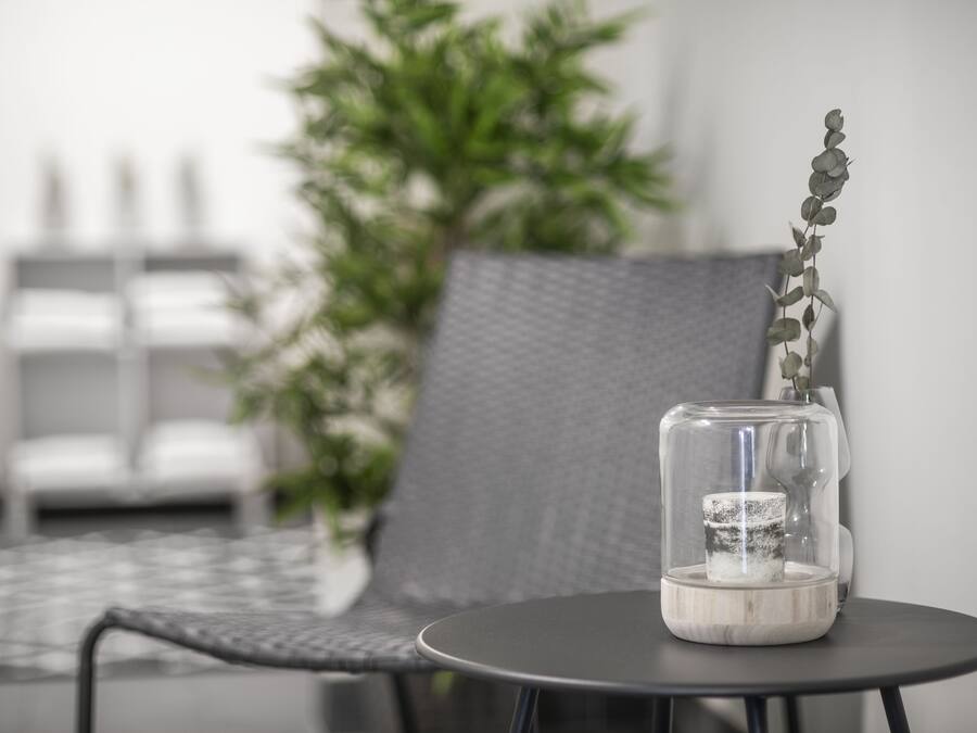 Quarto moderno com mesa redonda, estrutura de vidro com planta, cadeira cinza e estante ao fundo.
