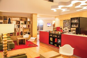 Um saguão de hotel moderno com um balcão de recepção vermelho, cadeiras brancas, uma estante de madeira e um tapete vermelho.