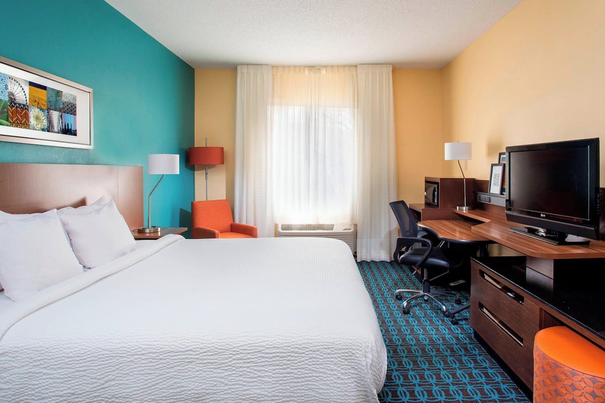 Quarto de hotel com uma cama grande, uma mesa com televisão, uma cadeira e uma luminária de mesa.
