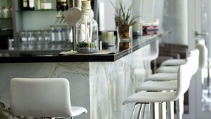 Uma área de bar com bancada de mármore, banquetas brancas e diversas garrafas e copos nas prateleiras.