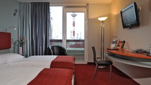 Quarto de hotel com duas camas, uma escrivaninha, uma cadeira, uma televisão e vista para um farol pela janela.