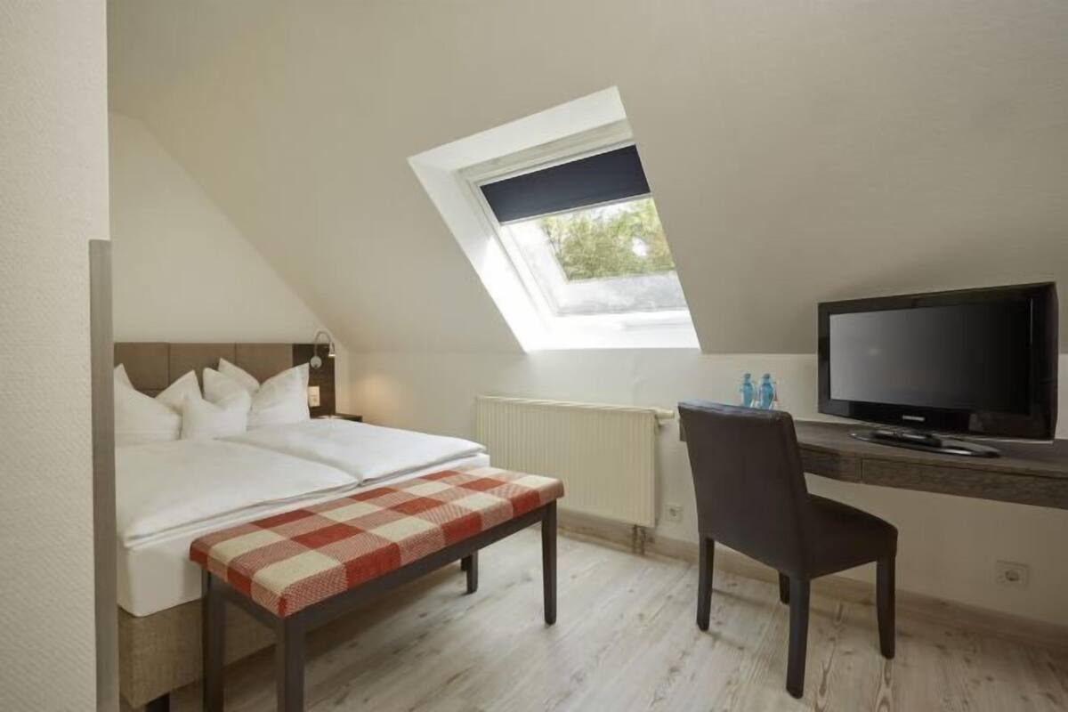 Quarto com claraboia, uma cama, um banco, uma escrivaninha com televisão e uma cadeira.