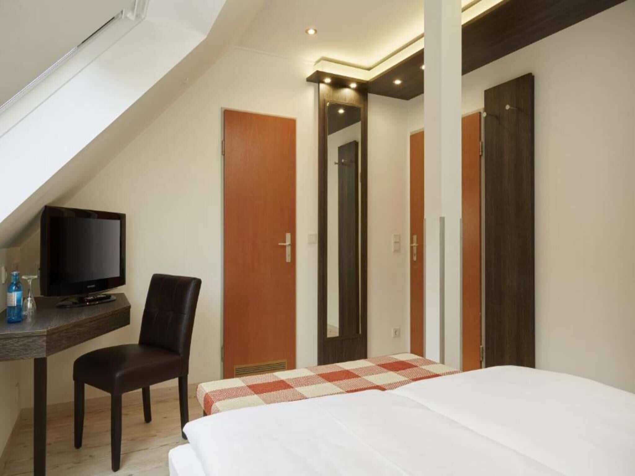 Quarto de hotel com cama, escrivaninha com televisão, uma cadeira e um guarda-roupa.