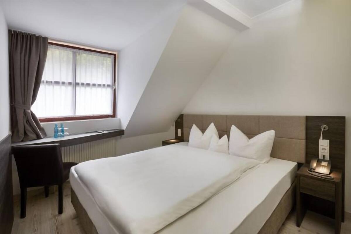 Quarto com uma cama grande, uma escrivaninha, uma cadeira, uma janela com cortinas e uma mesa de cabeceira com abajur.