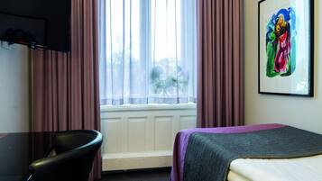 Quarto de hotel com cama, escrivaninha, cadeira, janela com cortinas e um quadro na parede.