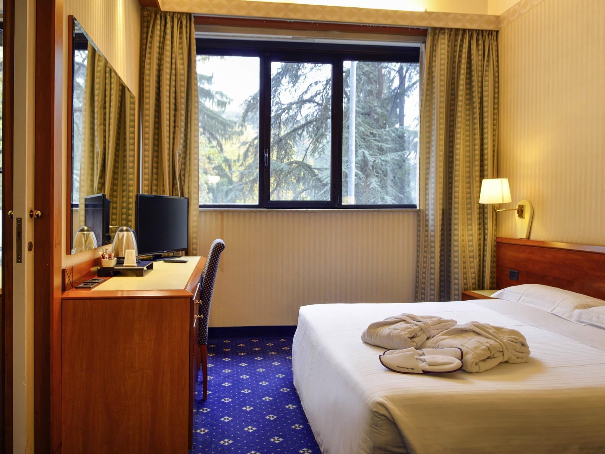 Quarto de hotel com uma cama, uma mesa com computador, uma cadeira, uma janela com cortinas e uma luminária.