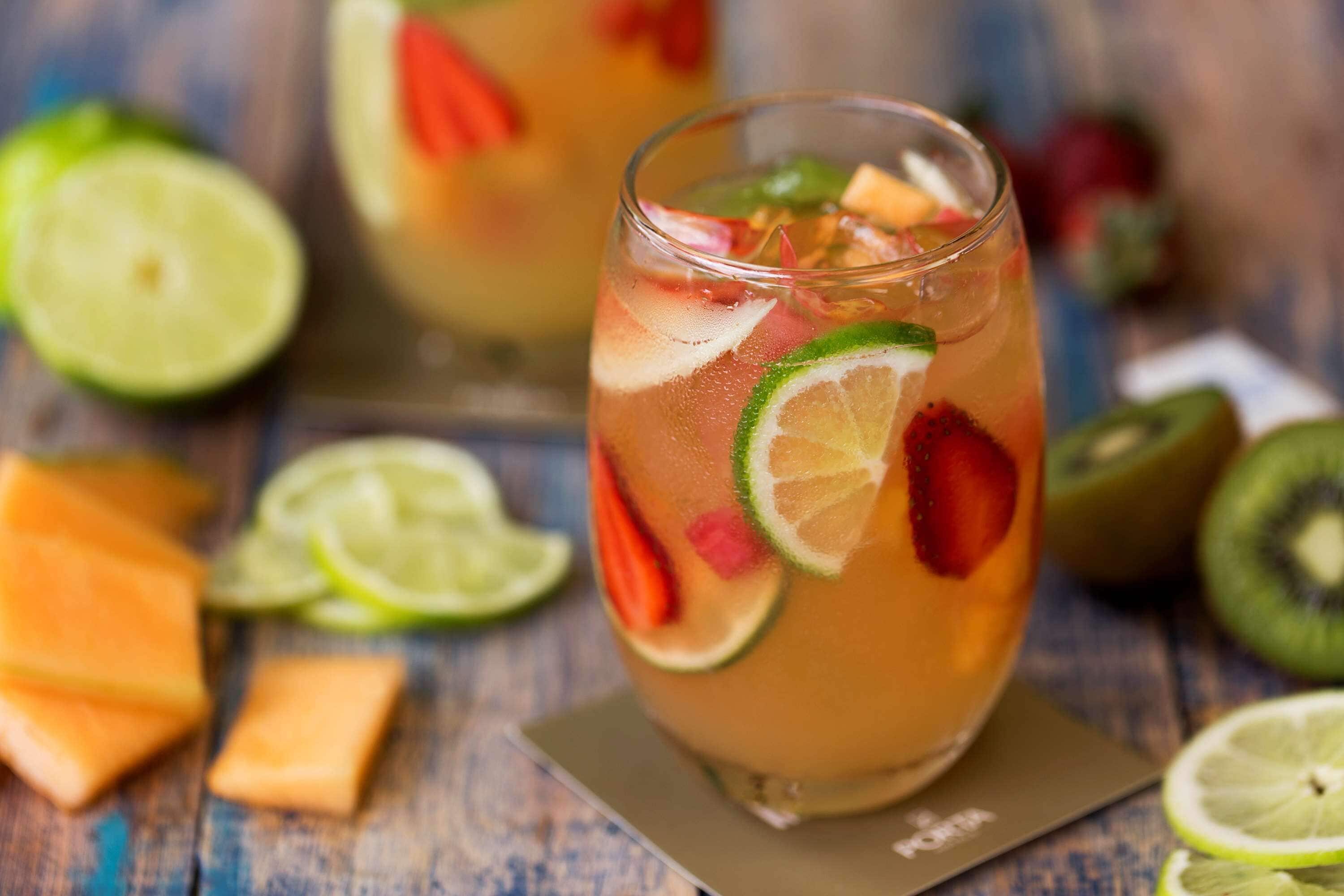 Um copo de bebida gelada com limão e fatias de frutas, cercado por frutas inteiras em uma superfície de madeira.