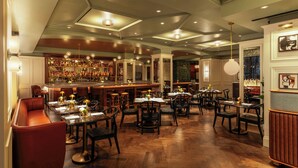 Restaurante com piso de madeira, várias mesas e cadeiras, área de bar com prateleiras de garrafas e iluminação pendente.