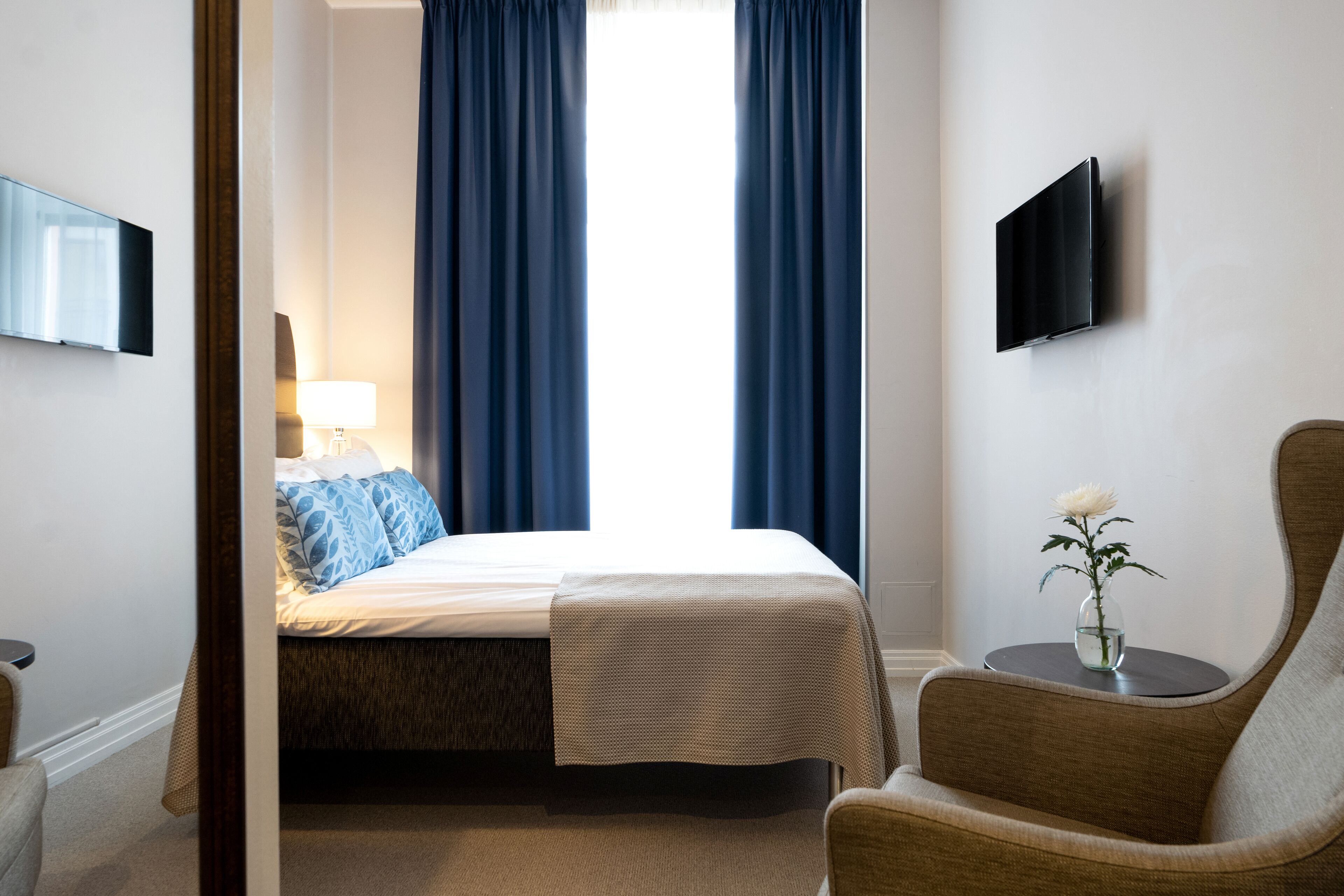 Quarto de hotel com cama, uma cadeira, televisão e um vaso com flores.
