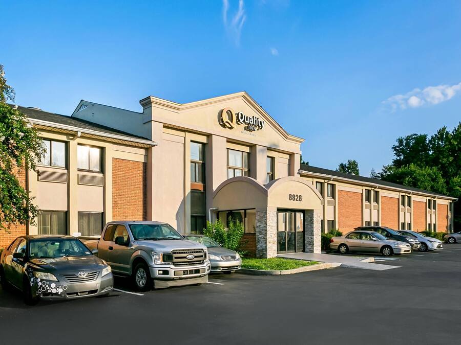 Um hotel com fachada bege, um letreiro grande de 'Quality Inn' e um estacionamento com vários carros.