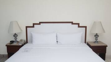 Cama bem arrumada com lençóis brancos, duas mesas de cabeceira com abajures e uma cabeceira de madeira.