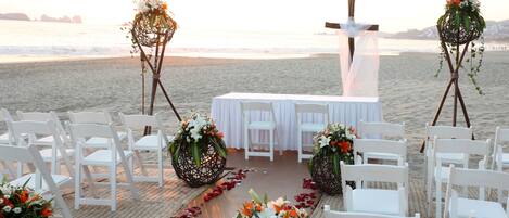 Decoração de casamento na praia, com cadeiras brancas e uma mesa adornada com flores e uma cruz.