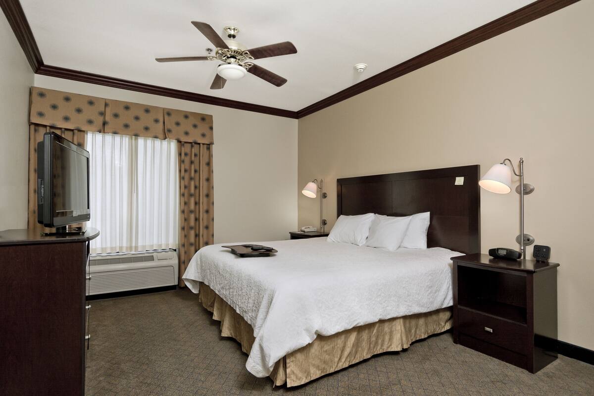 Quarto de hotel com uma cama, televisão, ventoinha no teto, mesa de cabeceira e abajur.