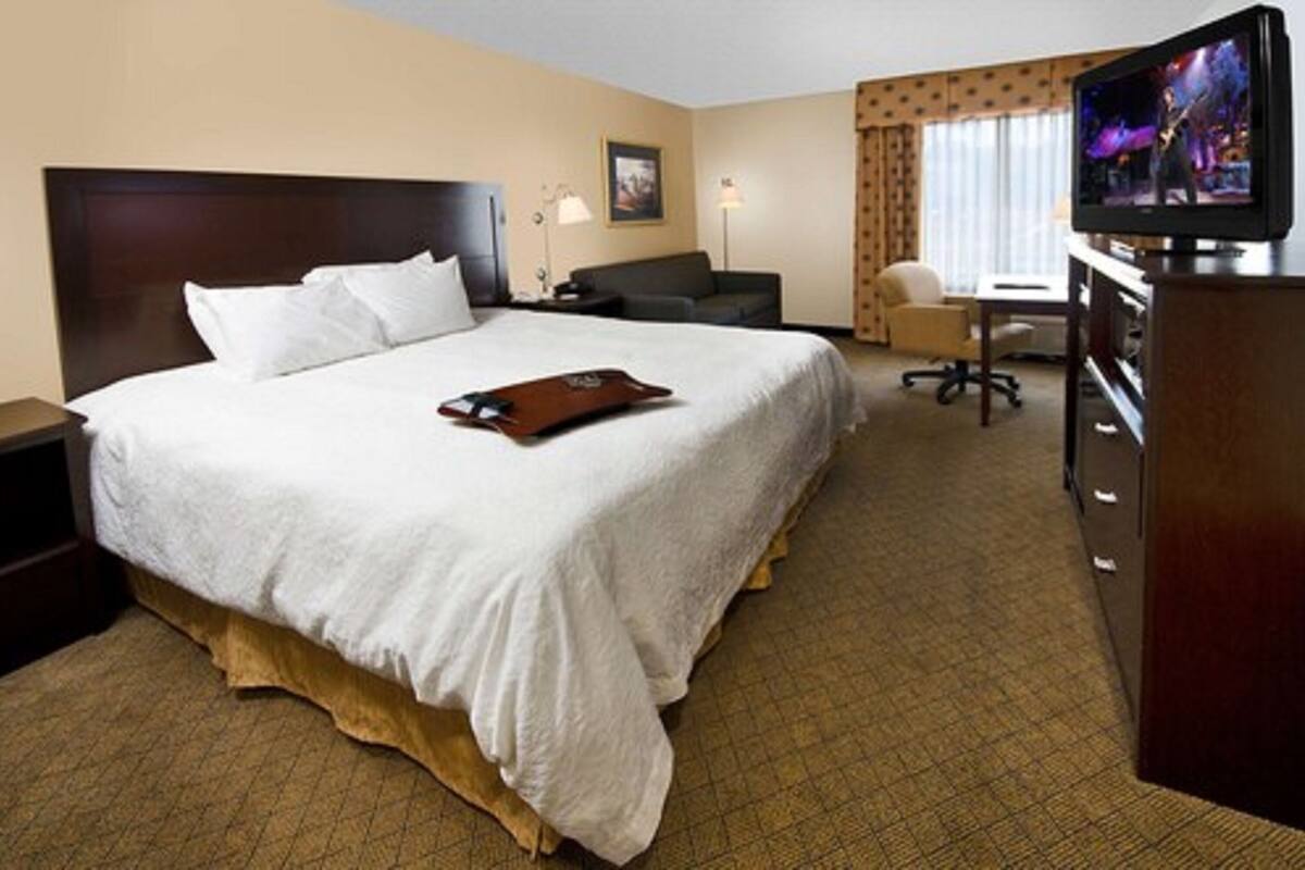 Quarto de hotel com uma cama grande, uma escrivaninha com televisão, uma cadeira e uma luminária de mesa.