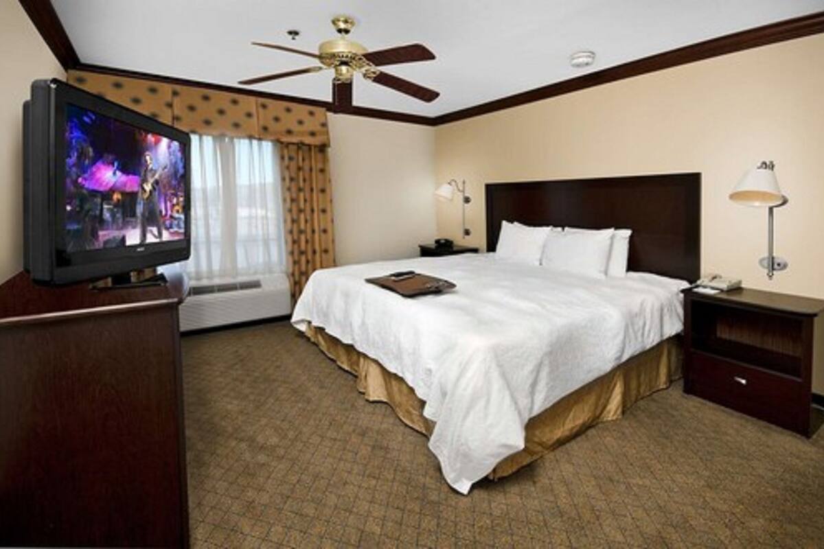Quarto de hotel com uma cama, televisão, ventoinha no teto e mesa de cabeceira.