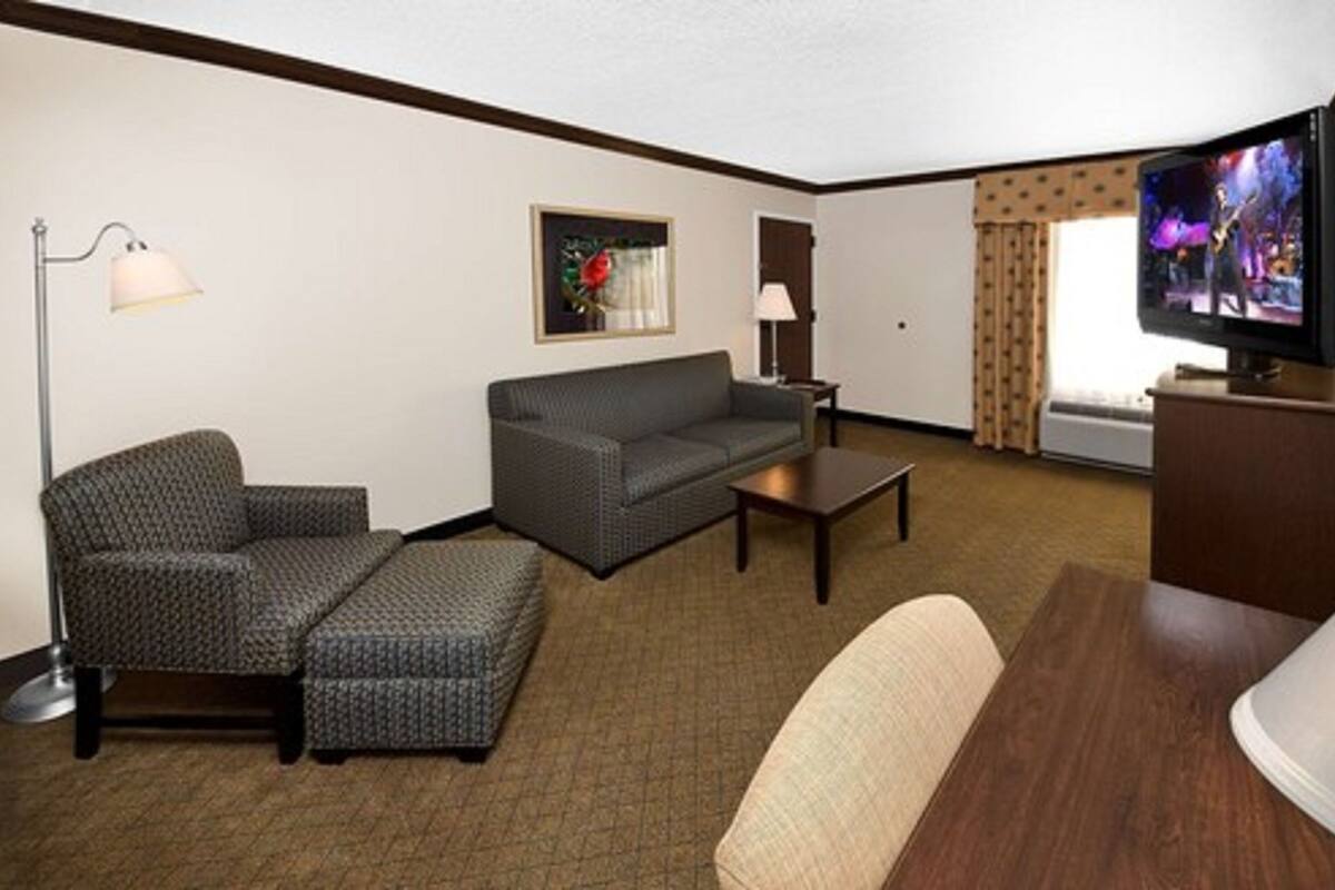 Quarto de hotel com sofá, banquinho, mesa de centro, abajur, televisão e um quadro pendurado na parede.