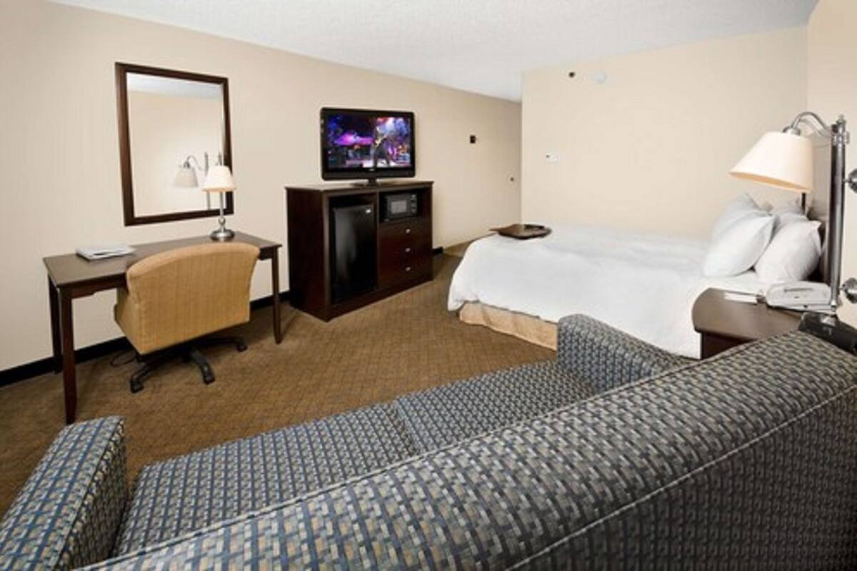 Quarto de hotel com cama, escrivaninha, cadeira, televisão e luminária.