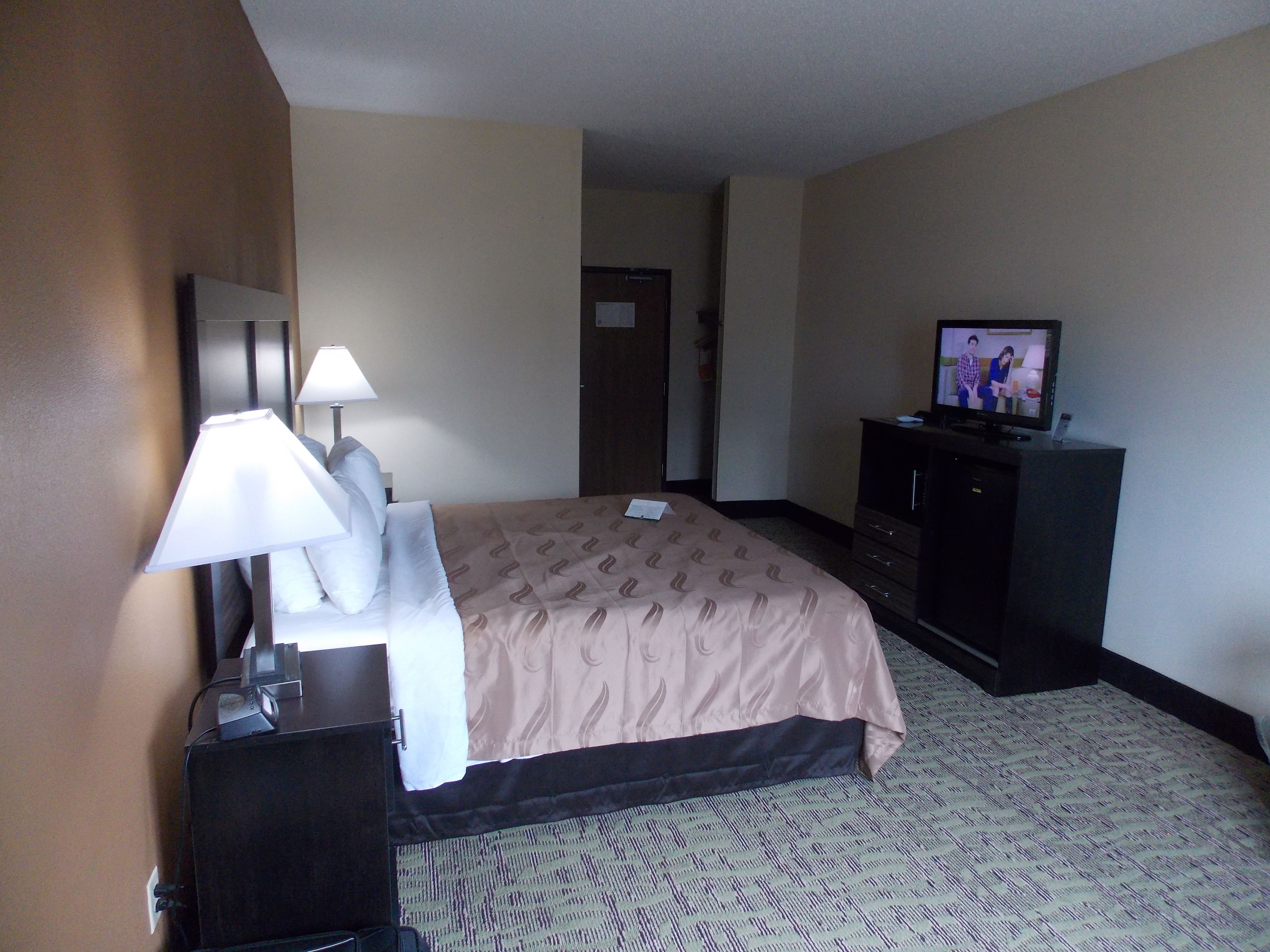Quarto de hotel com cama, escrivaninha com luminária, televisão e guarda-roupa.