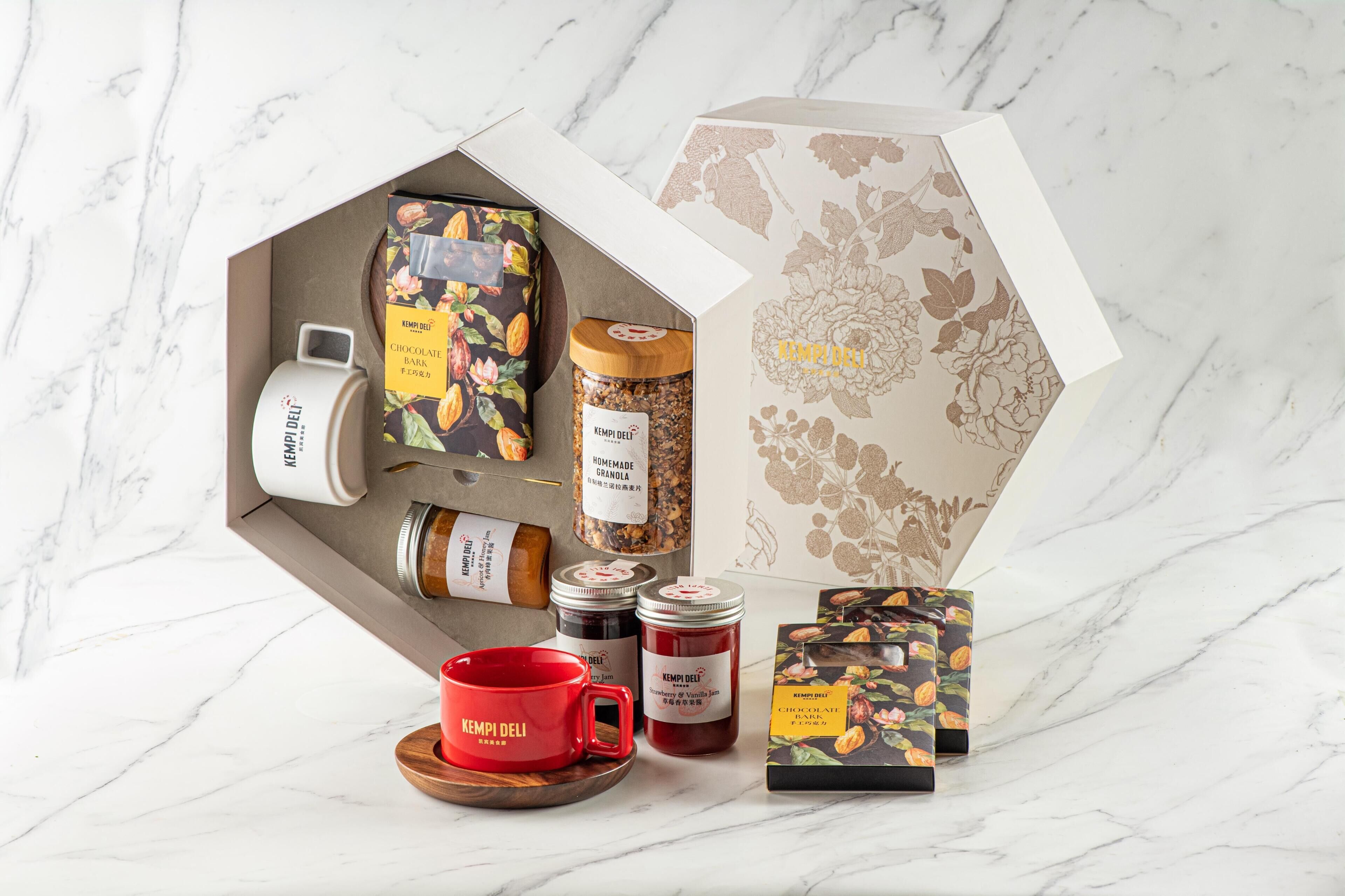 Uma caixa de presente com padrão floral, uma caneca vermelha e potes de granola e geleia caseiras.