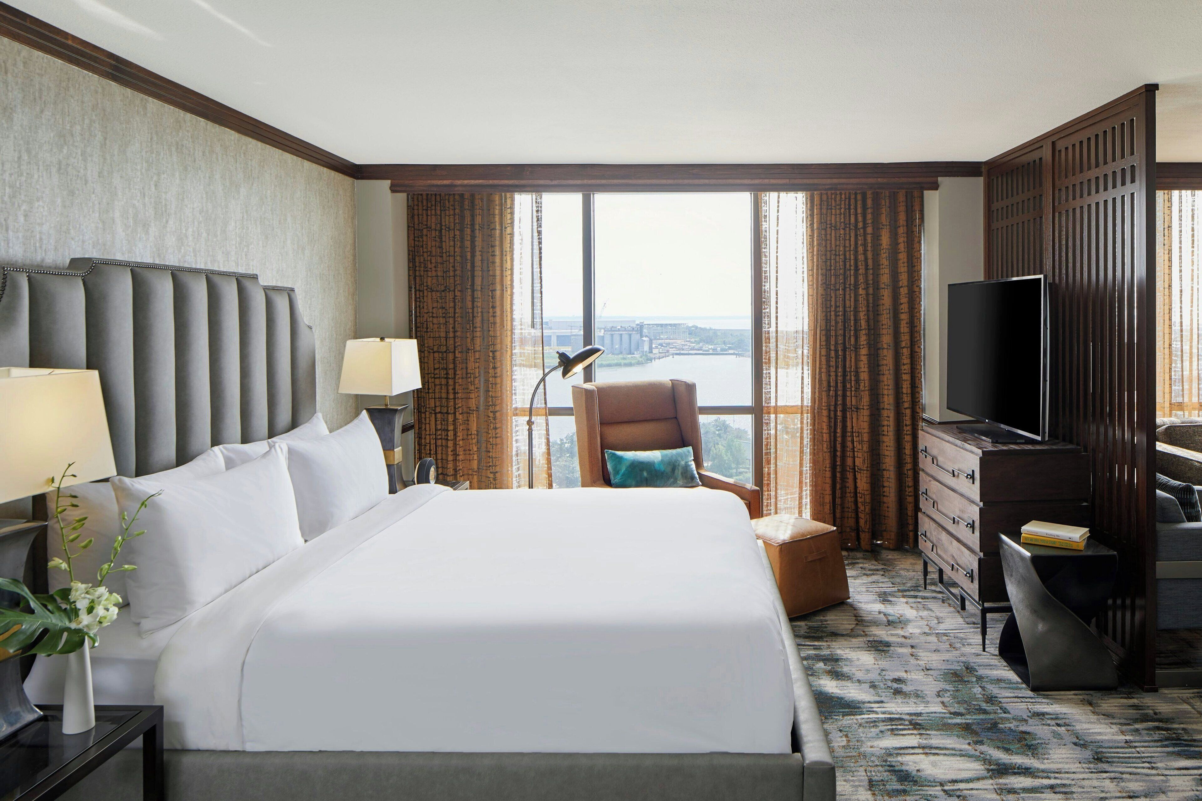 Quarto de hotel com uma cama grande, televisão, escrivaninha com abajur e vista para a cidade pelas janelas.