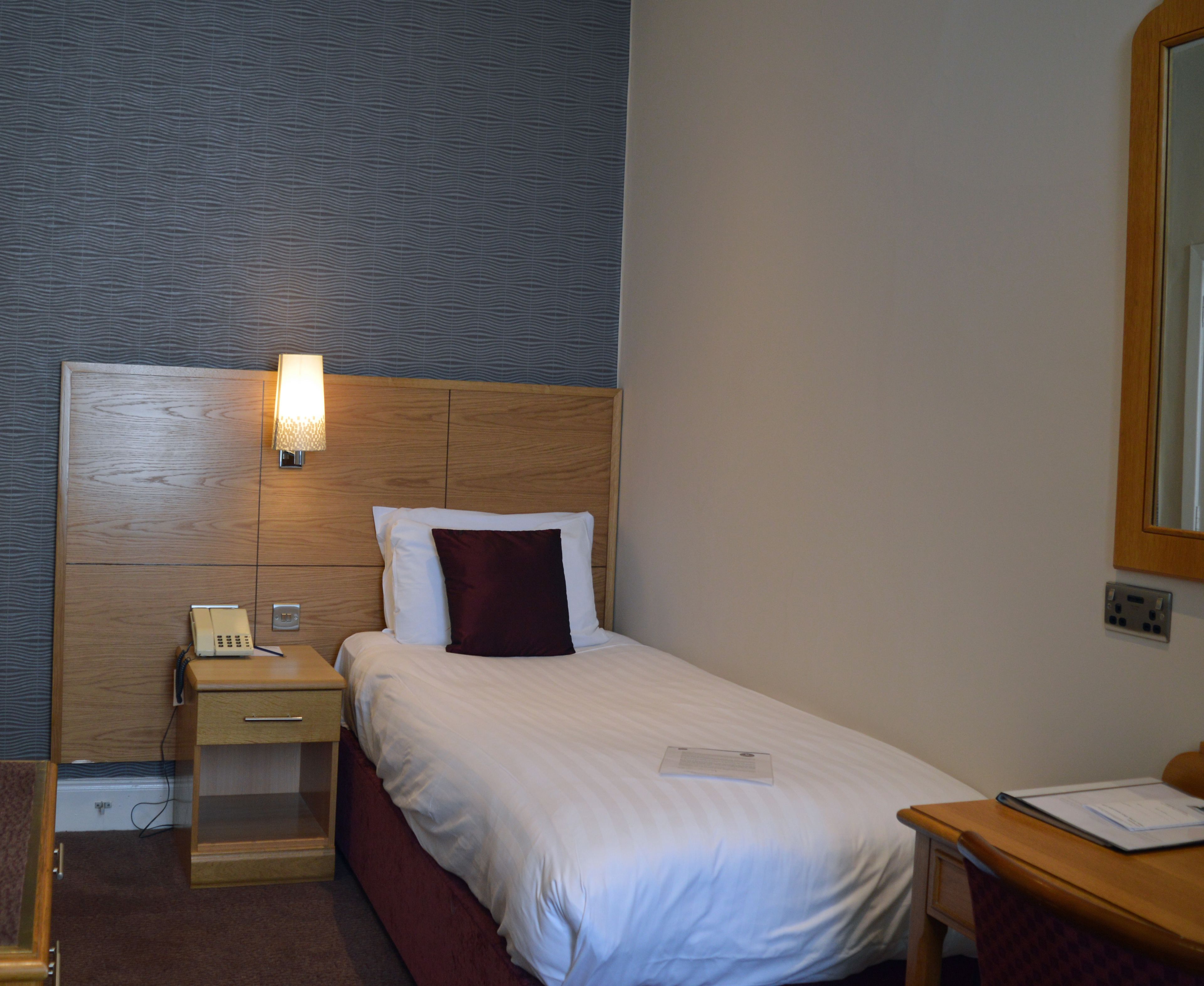 Quarto de hotel com cama, mesa de cabeceira com telefone, luminária na parede e espelho.