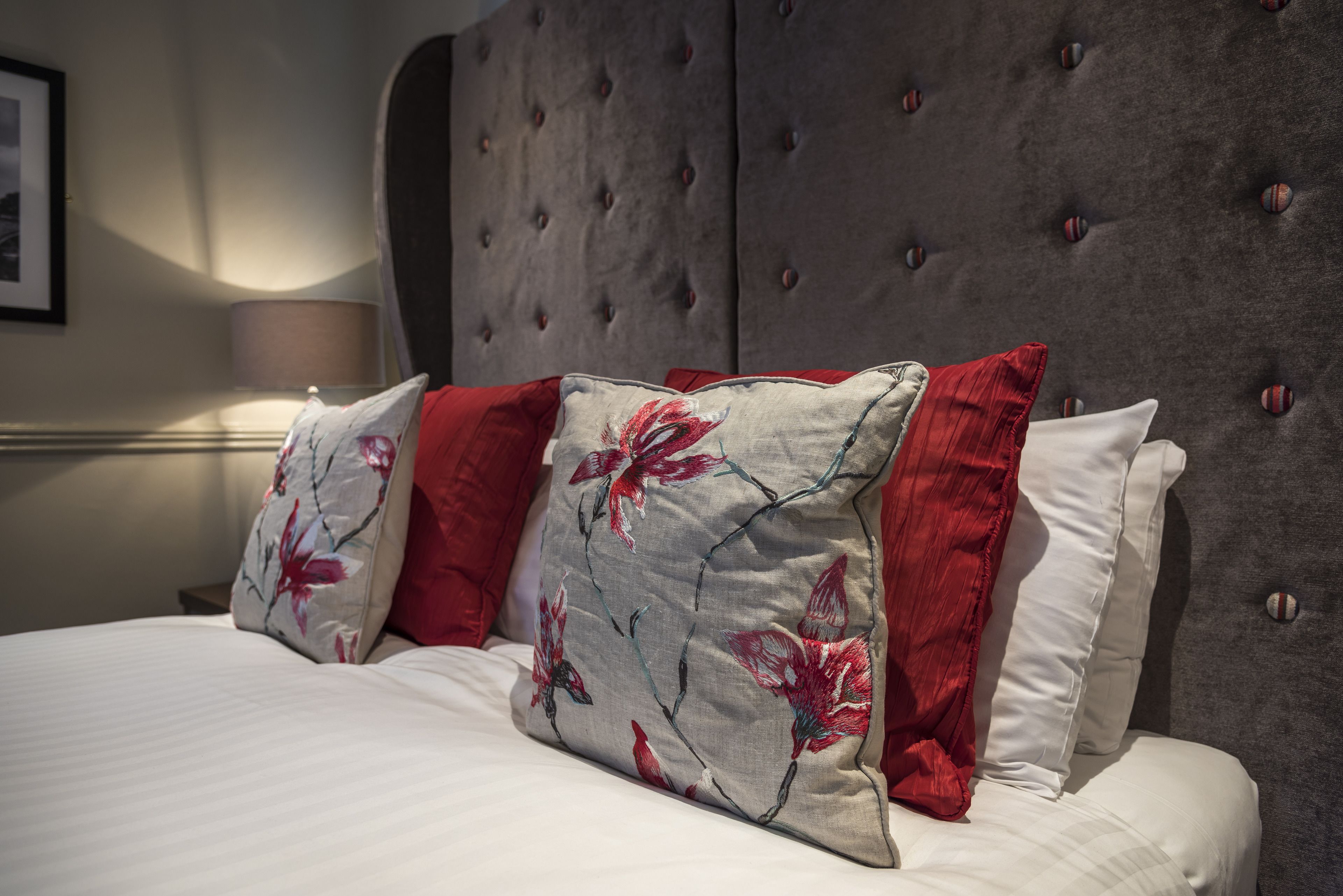 Cama com cabeceira estofada cinza, almofadas vermelhas e brancas e cobre cama branca.