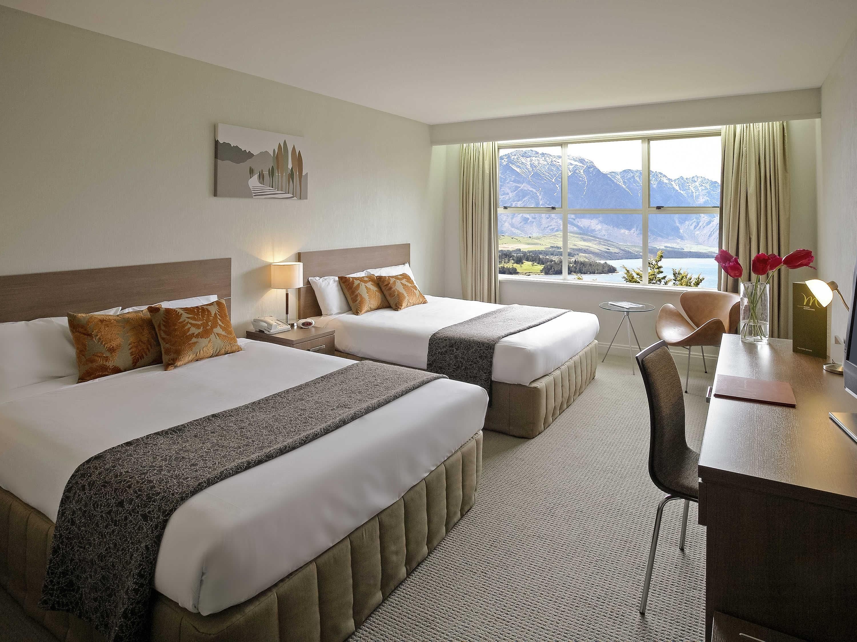 Quarto de hotel com duas camas, uma mesa, uma cadeira, vista para montanhas e um quadro na parede.