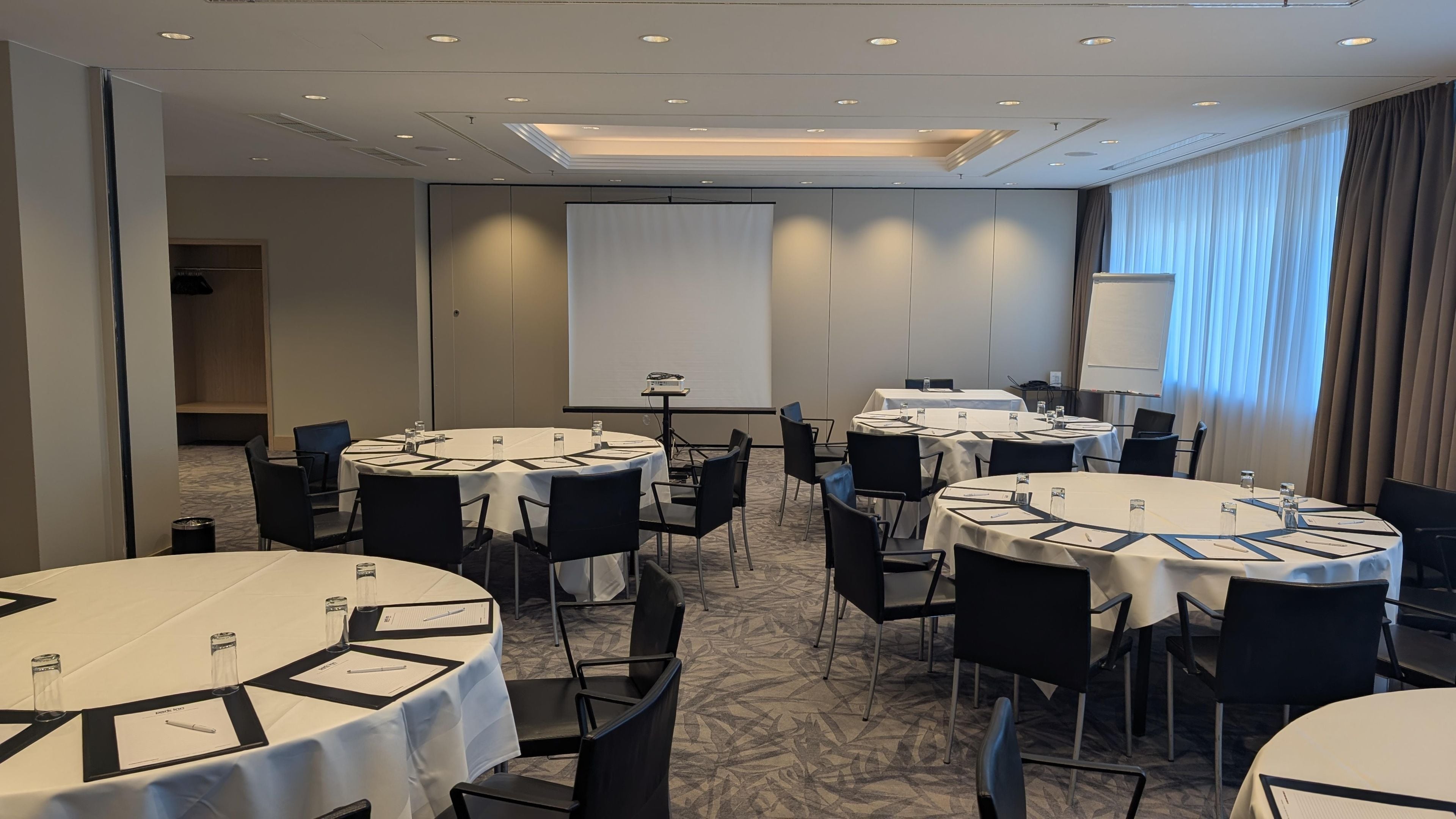 Sala de conferência com várias mesas redondas, cadeiras pretas, tela de projetor e quadro branco.