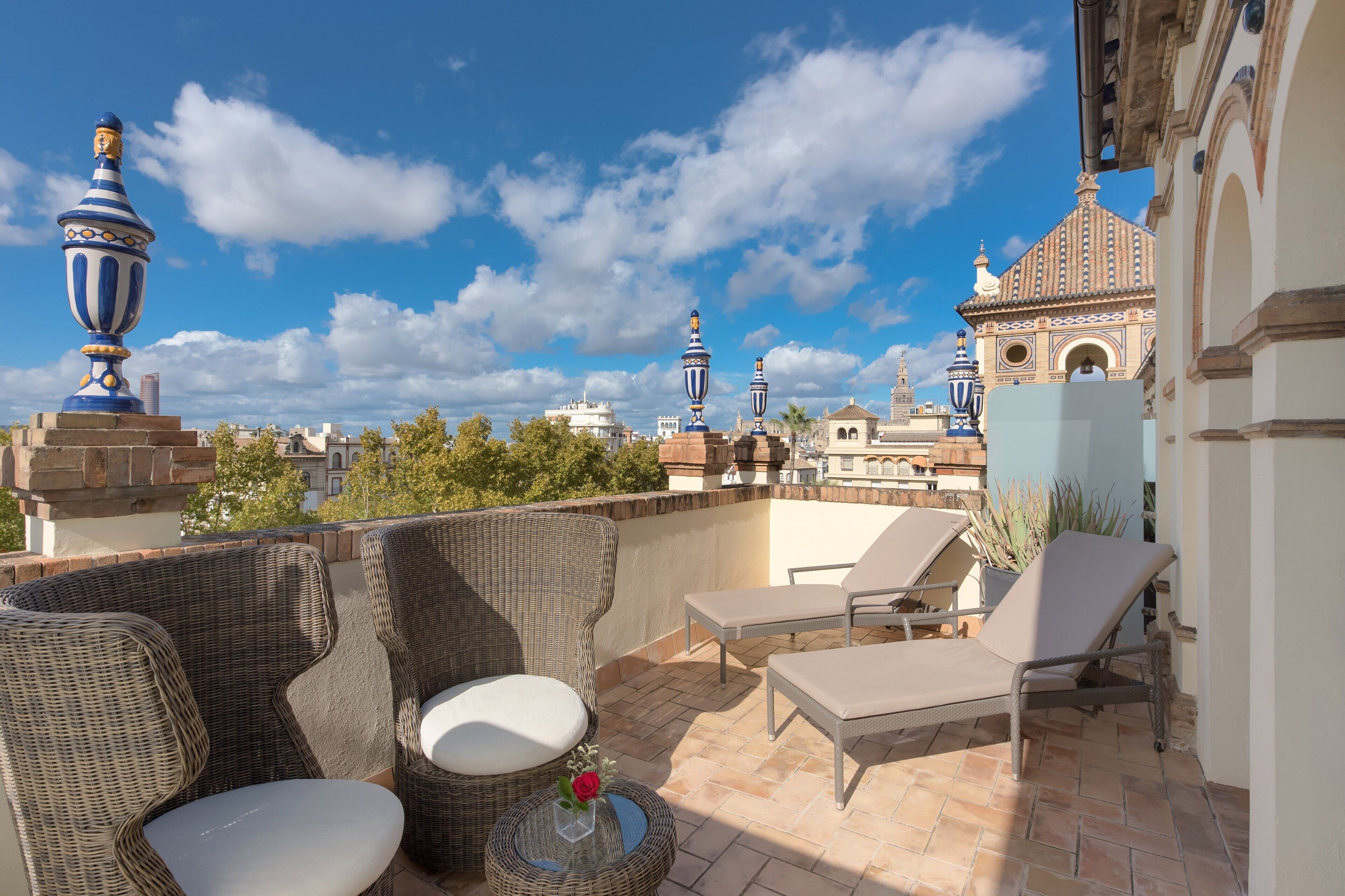 Terraço no telhado com móveis de vime, um vaso de flores e vista para edifícios históricos.