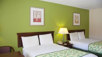 Quarto de hotel com duas camas, paredes verdes, pintura floral emoldurada, mesa de cabeceira com abajur e uma cadeira de madeira.